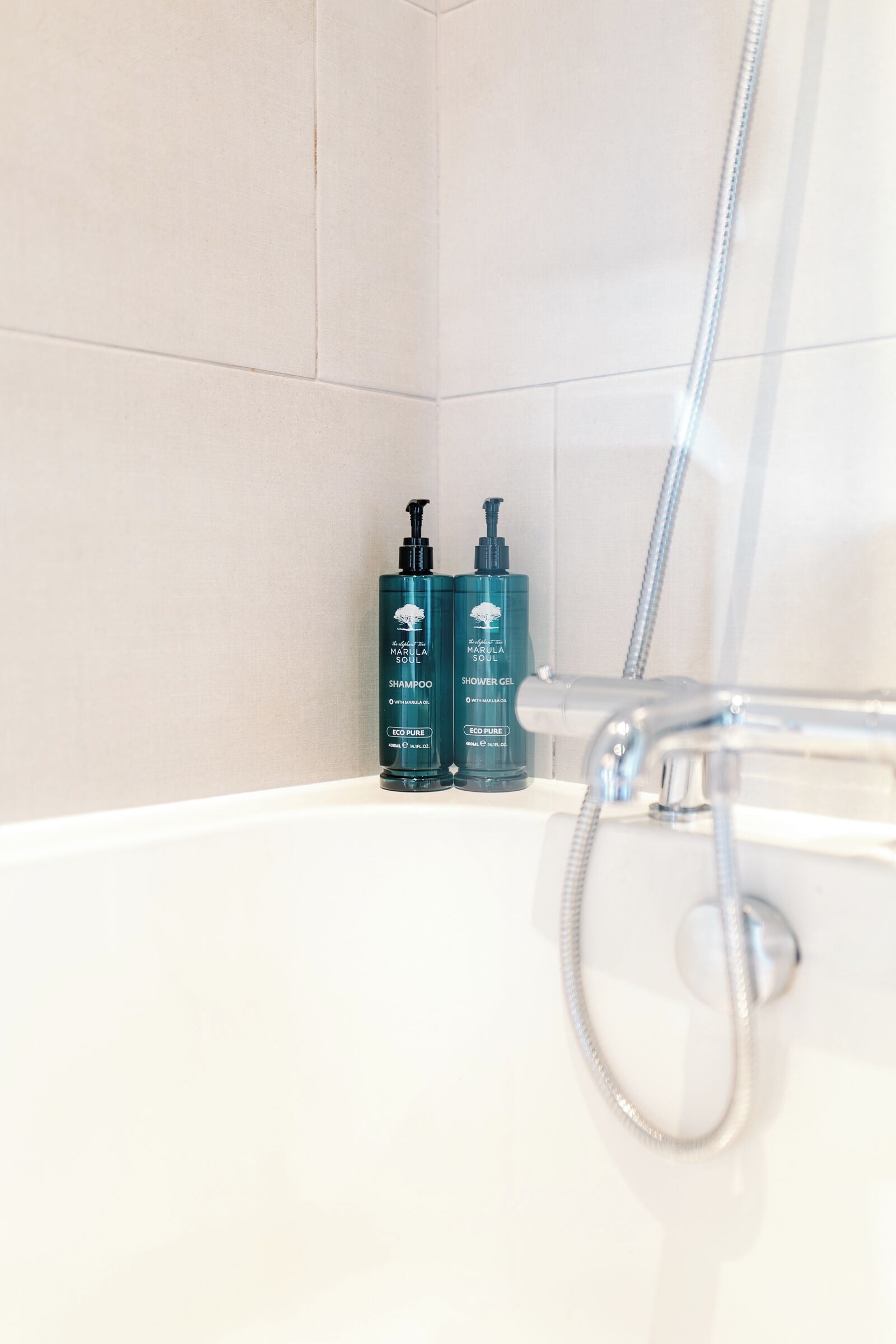 Casa de banho com chuveiro, dois frascos de shampoo e gel de banho numa prateleira, e um esguicho.