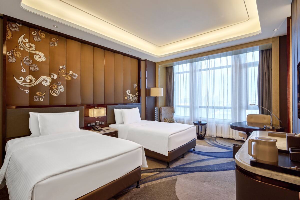 Quarto de hotel com duas camas, uma escrivaninha, uma cadeira, um abajur e uma janela grande com cortinas.