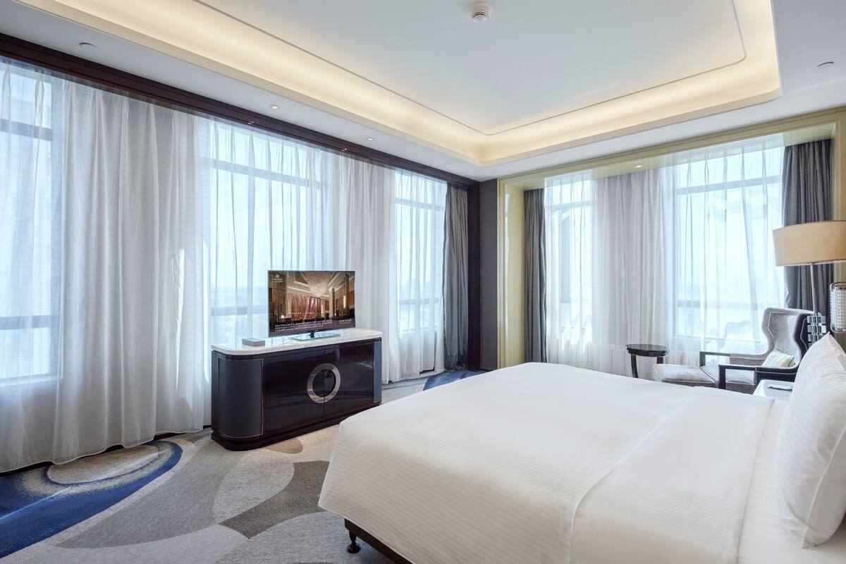 Quarto de hotel com uma cama grande, televisão em suporte, escrivaninha com cadeira e amplas janelas com cortinas.