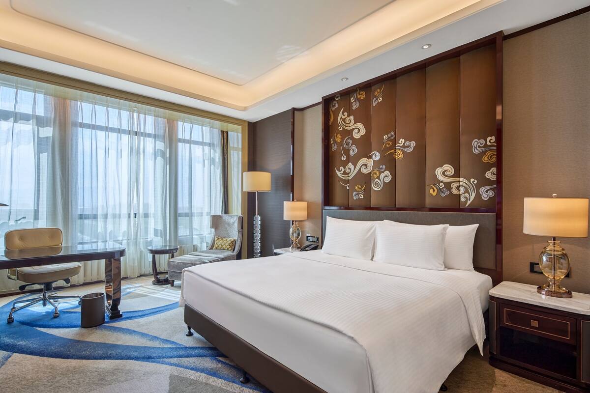 Quarto de hotel com uma cama grande, duas mesas de cabeceira com abajures, uma escrivaninha com cadeira e uma janela ampla com cortinas finas.