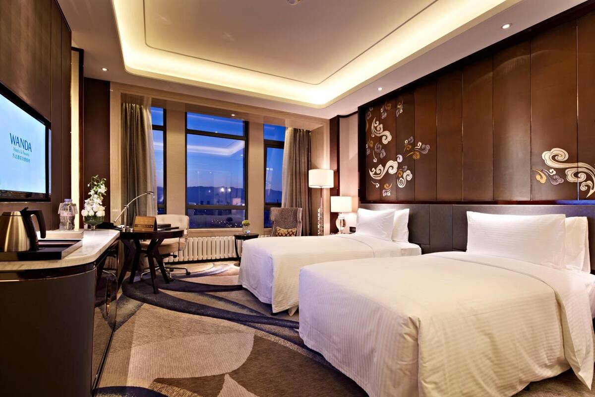 Quarto de hotel com duas camas, uma escrivaninha com computador, televisão e uma janela grande com vista para a cidade.