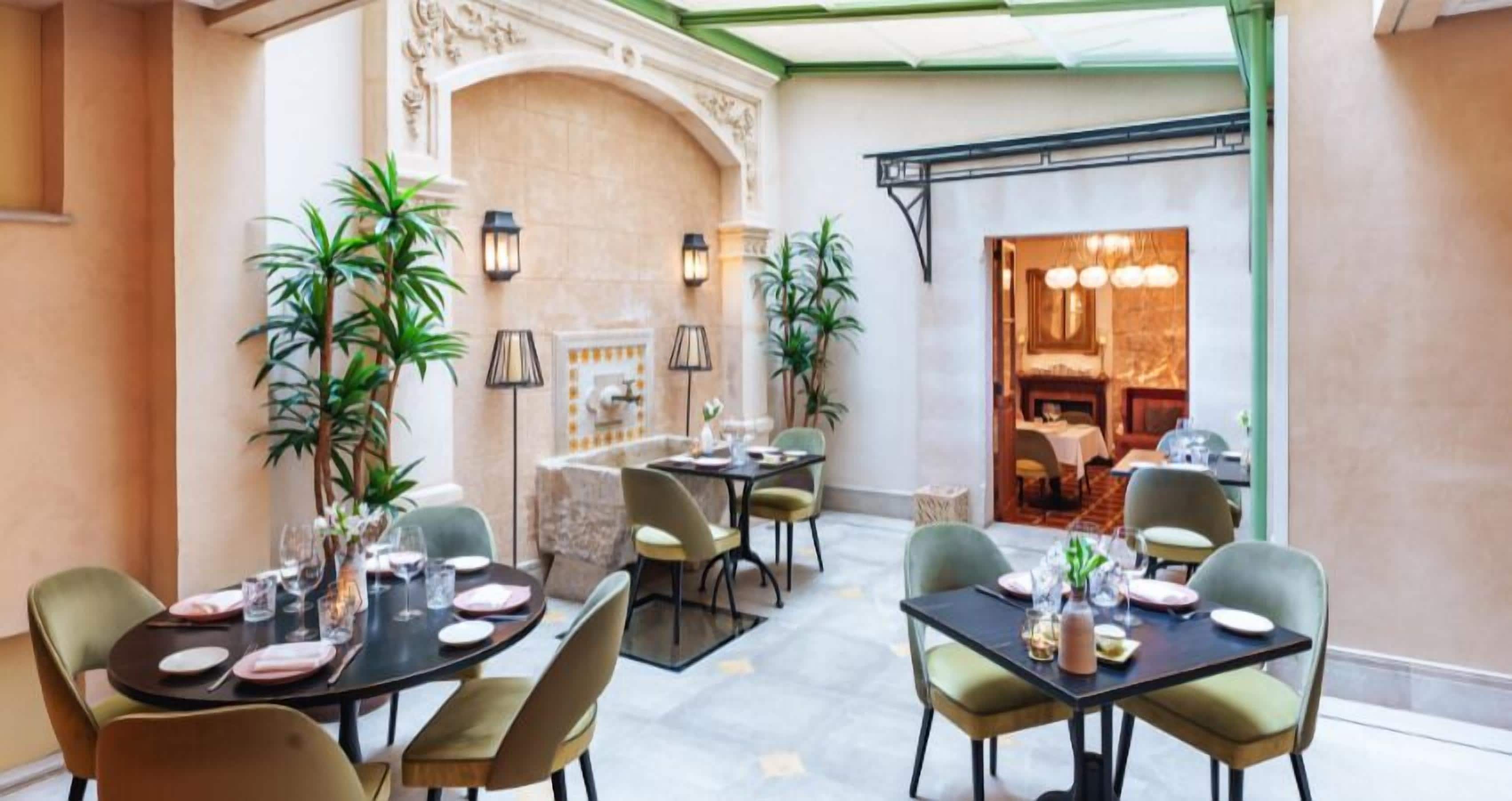 Uma área de refeições com mesas preparadas para quatro pessoas, rodeada por cadeiras, e vista para um restaurante através de uma abertura.