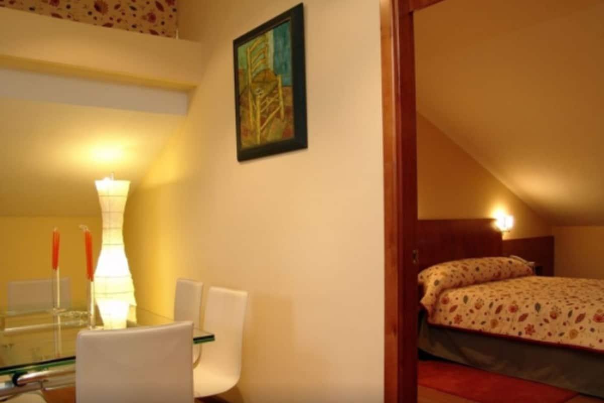 Quarto de hotel com uma cama, uma escrivaninha com cadeiras, um abajur e um quadro na parede.