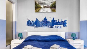 Uma cama com colcha azul, duas toalhas brancas e um mural na parede retratando a paisagem urbana de Roma.