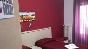 Quarto de hotel com três camas de solteiro, parede vermelha, quadro com vista urbana e placa indicando o tipo de quarto.