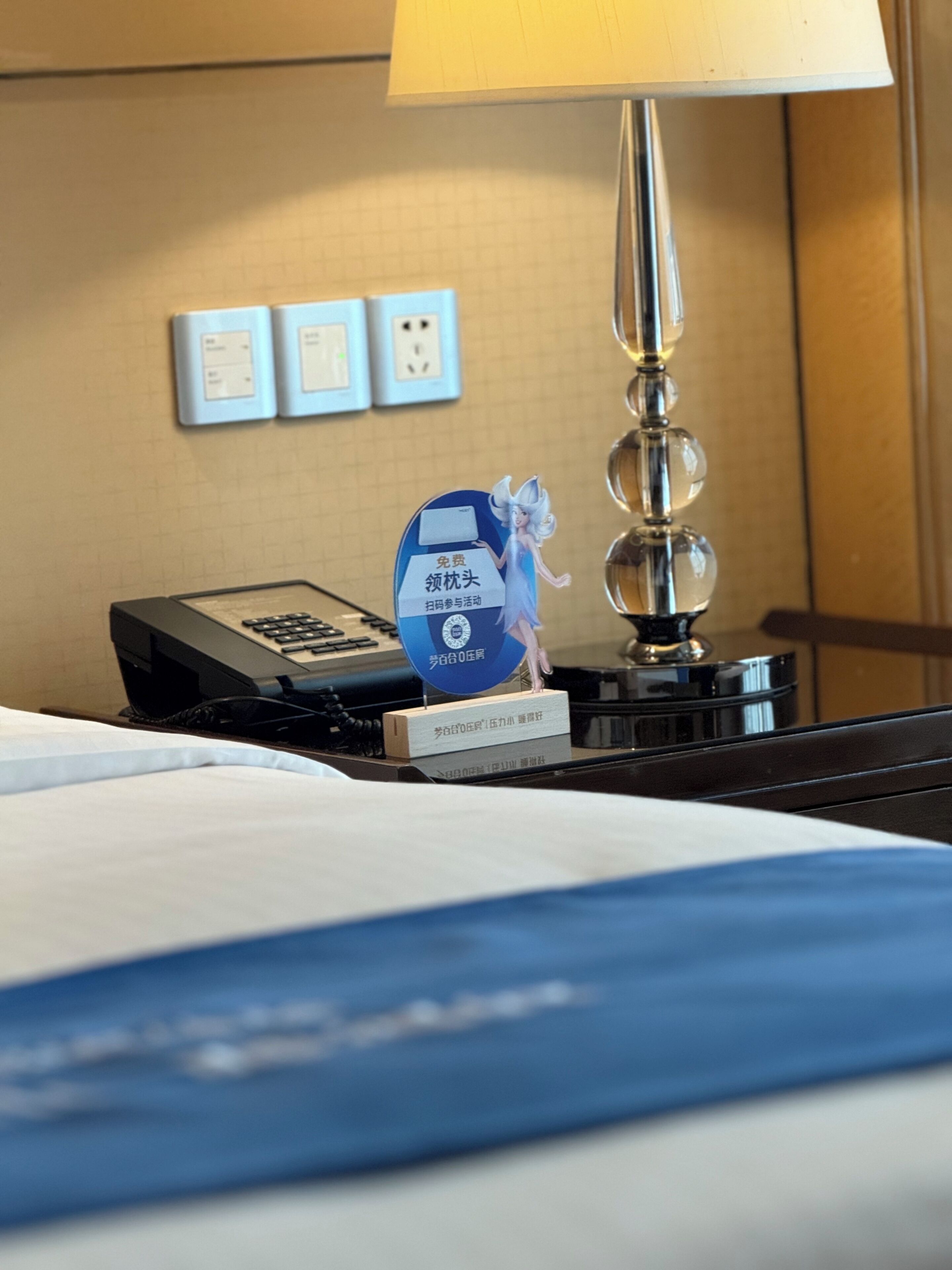 Quarto de hotel com uma cama, uma luminária, um telefone e uma tomada na parede.