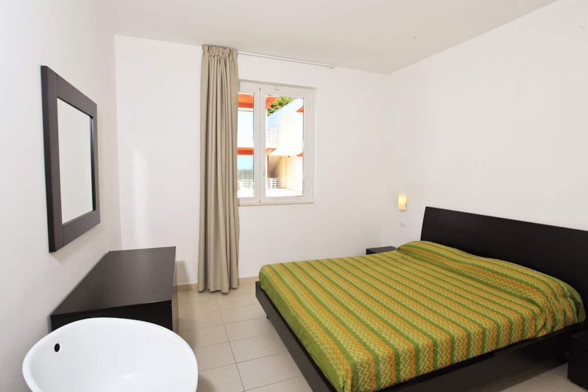 Um quarto com uma cama, uma janela com cortinas, uma mesa de cabeceira e uma pia branca.