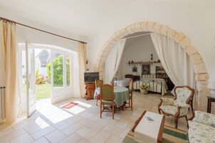 Uma sala de jantar com mesa e cadeiras, uma televisão e vista para o jardim através de uma porta aberta.