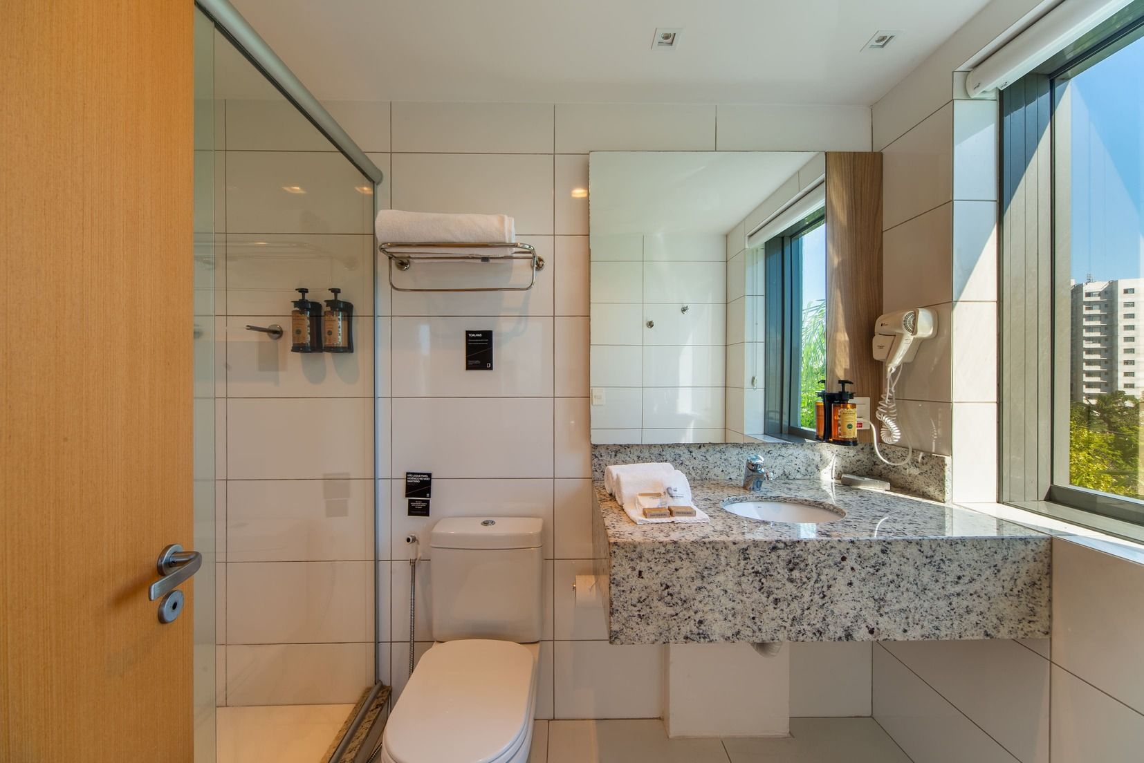 Casa de banho moderna com cabine de duche em vidro, sanita, lavatório com bancada e secador de cabelo fixado na parede.