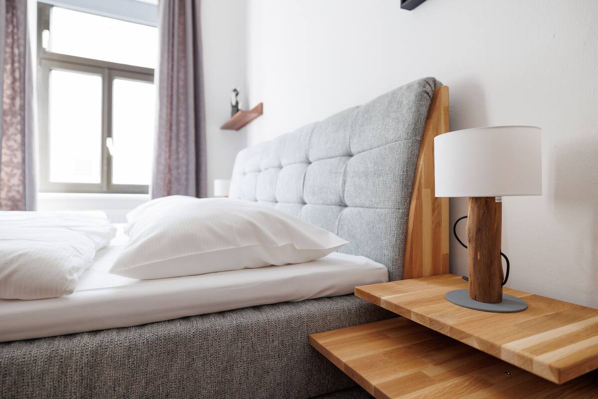 Uma cama bem arrumada com lençóis e almofadas brancas, uma mesa de cabeceira de madeira com uma luminária e uma janela com cortinas.