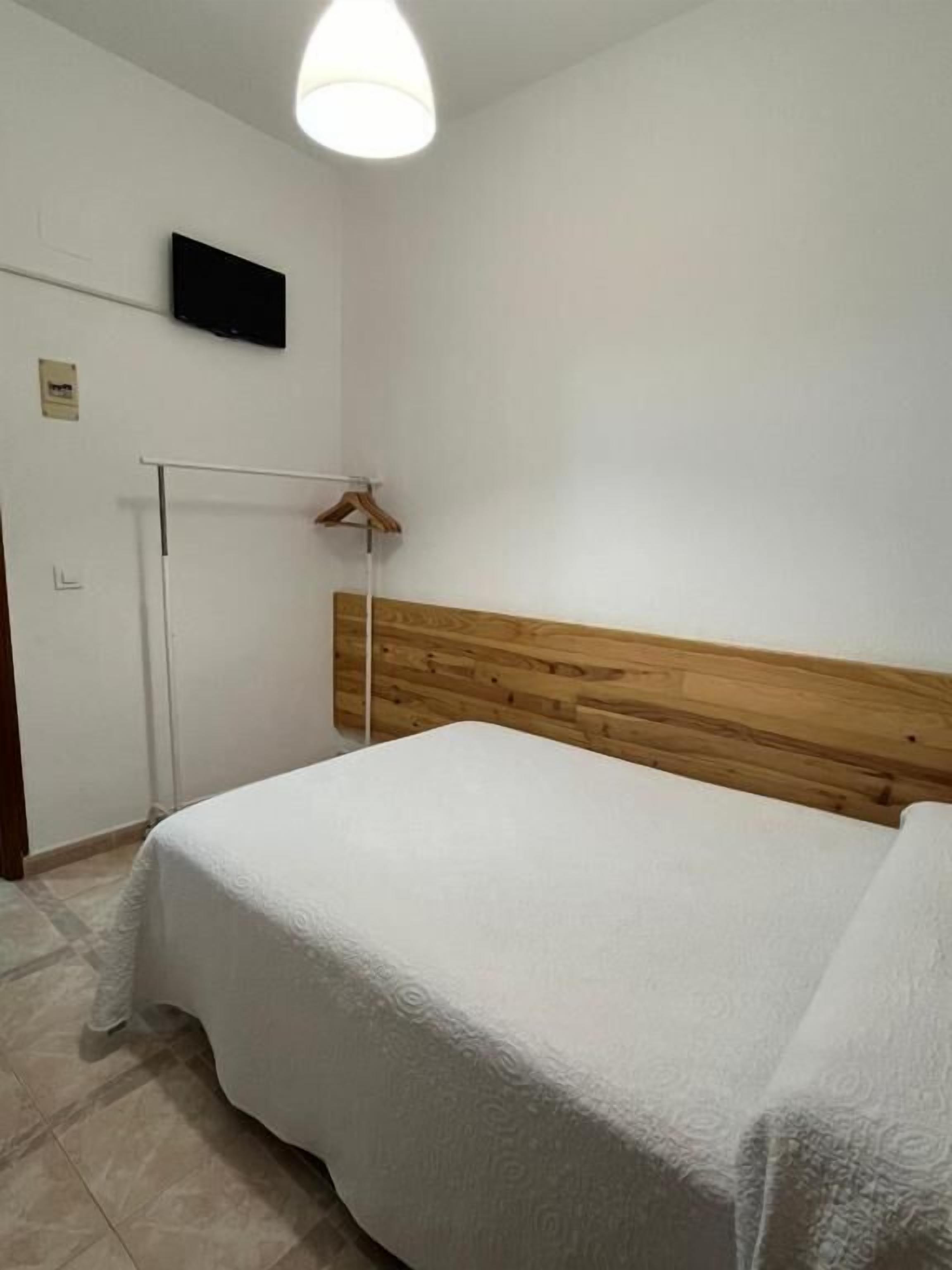 Quarto com cabeceira de madeira, colcha branca, cabideiro e televisão na parede.