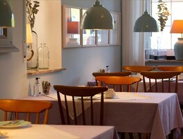 Espaço de jantar com mesas preparadas para quatro pessoas, cadeiras de madeira e iluminação pendente.