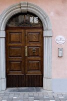 Uma porta dupla de madeira com puxador de metal, emoldurada por uma estrutura de pedra em arco, contra uma parede rosa com uma placa que diz