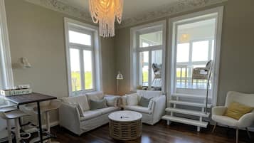 Ampla sala de estar com um grande candelabro, sofás brancos, uma mesa de centro e um espaço de jantar com cadeiras de vime.