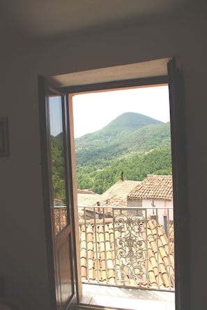 Vista de um quarto com uma grande janela, mostrando uma paisagem montanhosa e telhados de terracota.
