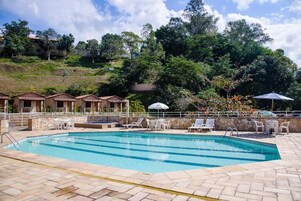 Piscina com espreguiçadeiras e guarda-sóis, rodeada por um muro de pedra e vegetação.