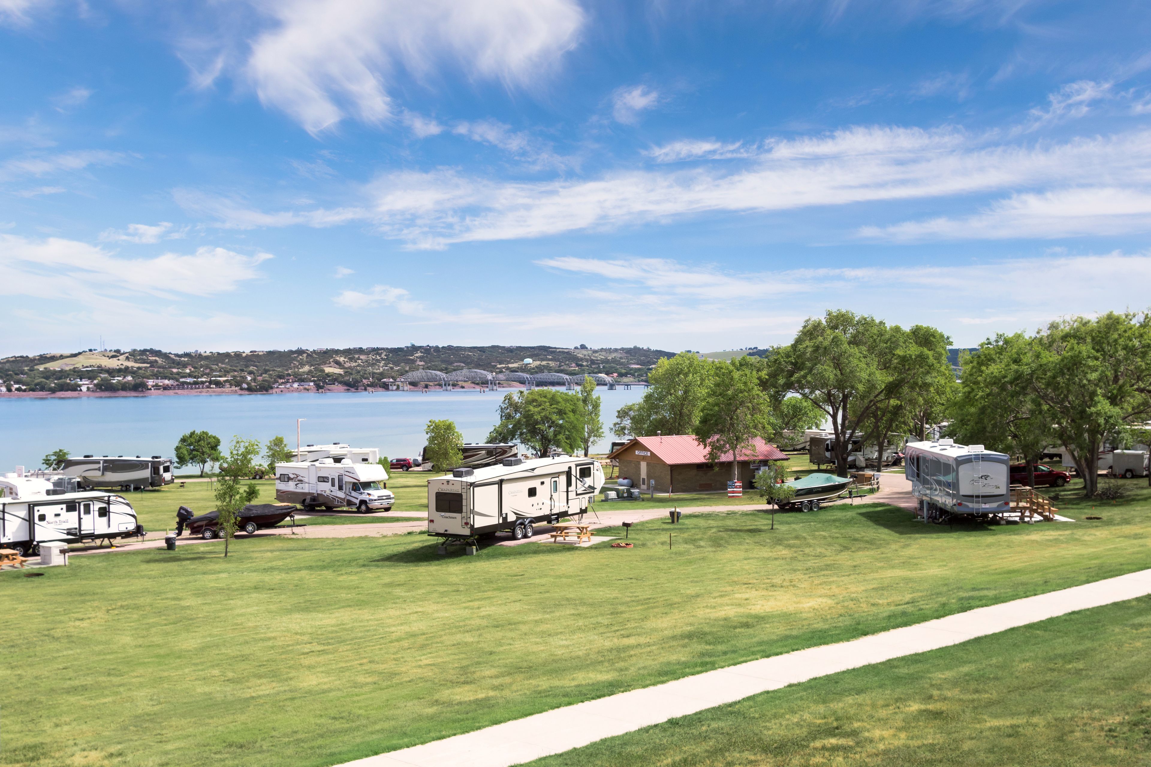 Um parque de campismo com veículos recreativos estacionados num campo relvado, um pequeno edifício e um lago ao fundo.