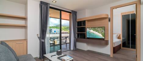 Quarto de hotel moderno com sofá, uma mesinha, televisão montada na parede, varanda com vista para a cidade e porta deslizante que dá para um quarto.