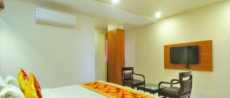 Quarto de hotel com uma cama, uma secretária com duas cadeiras, um ventilador de teto, uma televisão de tela plana na parede e um aparelho de ar condicionado.