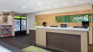 Uma área de recepção de hotel com design moderno, incluindo um balcão de recepção, um quiosque digital e uma máquina de vendas automática.