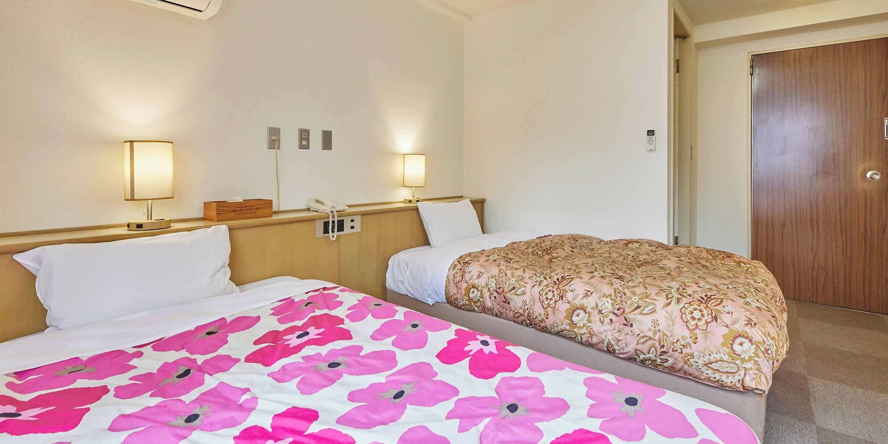 Quarto de hotel com duas camas, cada uma com colcha de padrão diferente. Há luminárias laterais e um telefone na mesa de cabeceira.