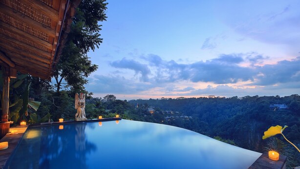 Uma piscina infinita com velas, cercada por vegetação exuberante e vista para uma aldeia distante.