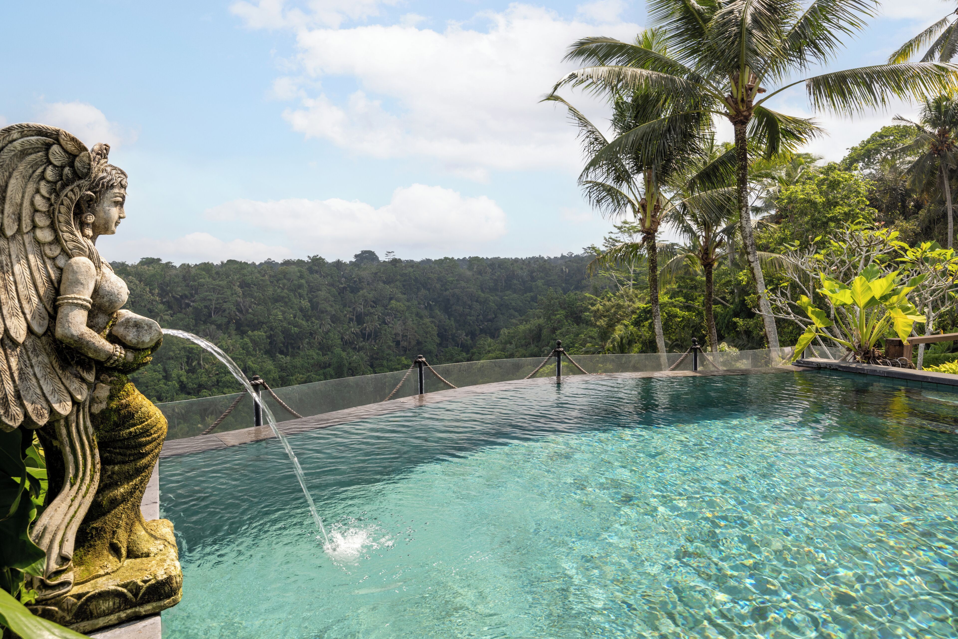 Vista clara de uma piscina com uma estátua à esquerda, rodeada por vegetação exuberante e palmeiras.