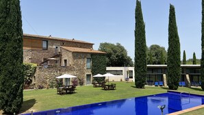 Um edifício de pedra com telhado de azulejos, piscina e altas árvores de cipreste.