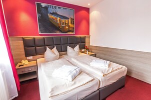 Quarto de hotel com cama de casal, duas mesas de cabeceira, criado-mudo e televisão instalada na parede.