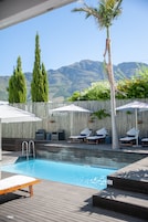Área de piscina com espreguiçadeiras e guarda-sóis, cercada por uma cerca de madeira e palmeiras, com montanhas ao fundo.