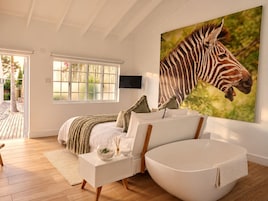 Quarto com uma cama grande, uma mesa de cabeceira branca com uma tigela de vidro, uma banheira branca espaçosa e uma pintura de zebra na parede.