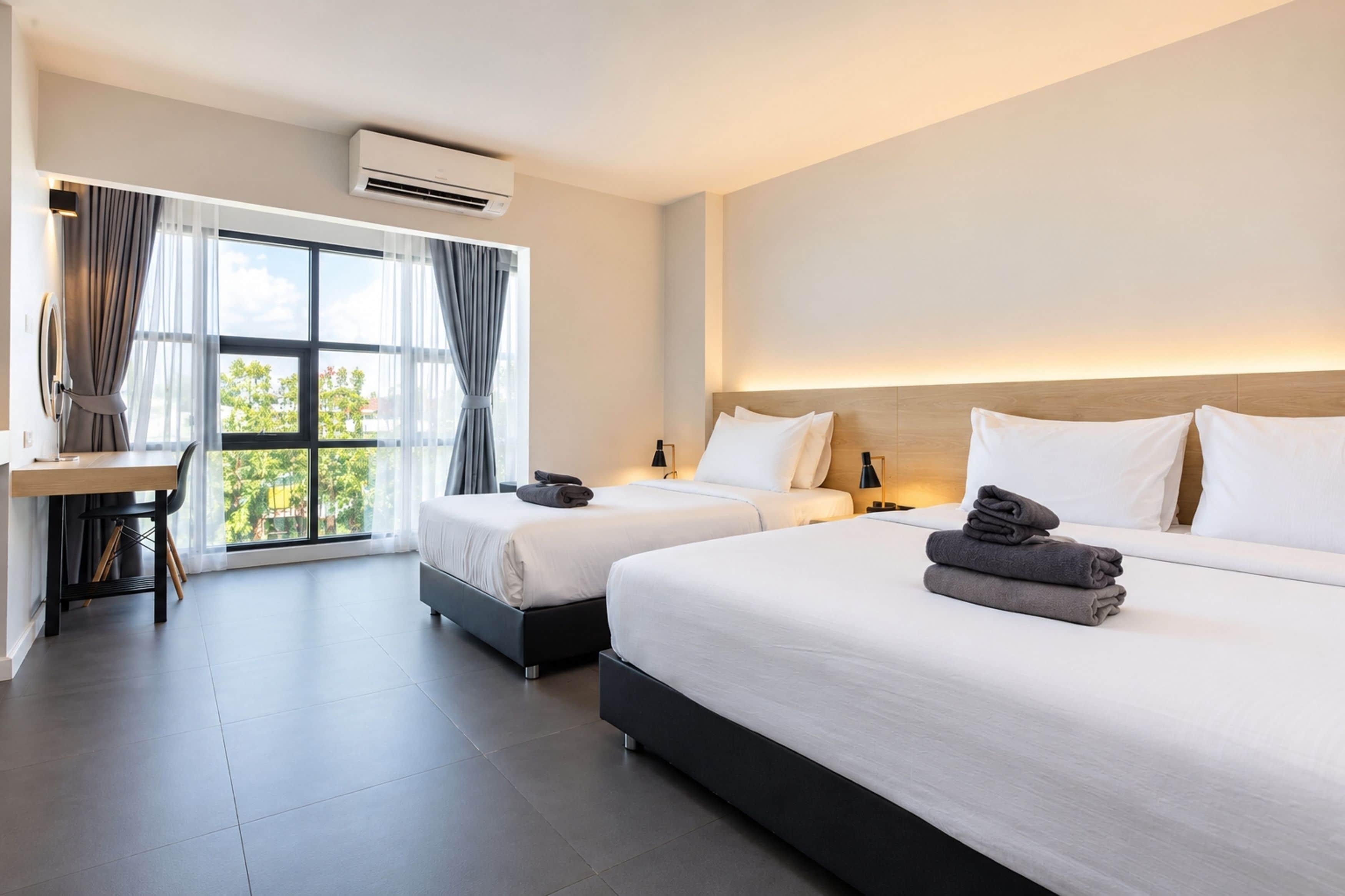 Quarto de hotel com duas camas, uma mesa de trabalho e uma janela grande com cortinas.
