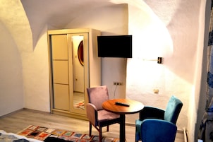 Um quarto pequeno e bem iluminado com uma mesa redonda, duas cadeiras, uma televisão fixada na parede e uma porta que dá para outro cômodo.