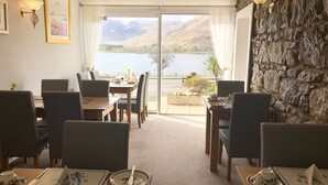 Uma sala de jantar com mesas preparadas para dois, cadeiras e vista para um lago e montanhas.