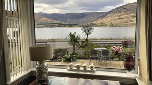 Vista da janela sobre um lago com montanhas, uma pequena mesa com abajur e plantas em vasos.