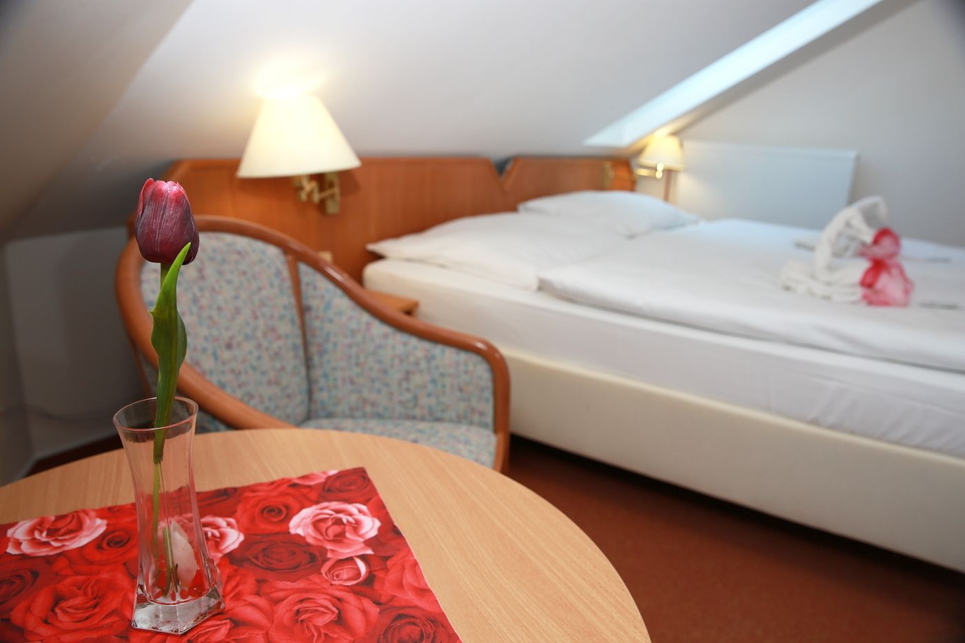 Quarto de hotel com cama, mesa de cabeceira, abajur e um vaso com uma única flor.