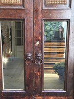 Uma porta de madeira com dois chocalhos em forma de cabeça de le ão e um painel de vidro refletindo uma pessoa e uma parede de tijolos.