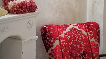 Canto de um quarto com parede branca, um armário branco com detalhes florais entalhados e uma cadeira com padrão floral vermelho.