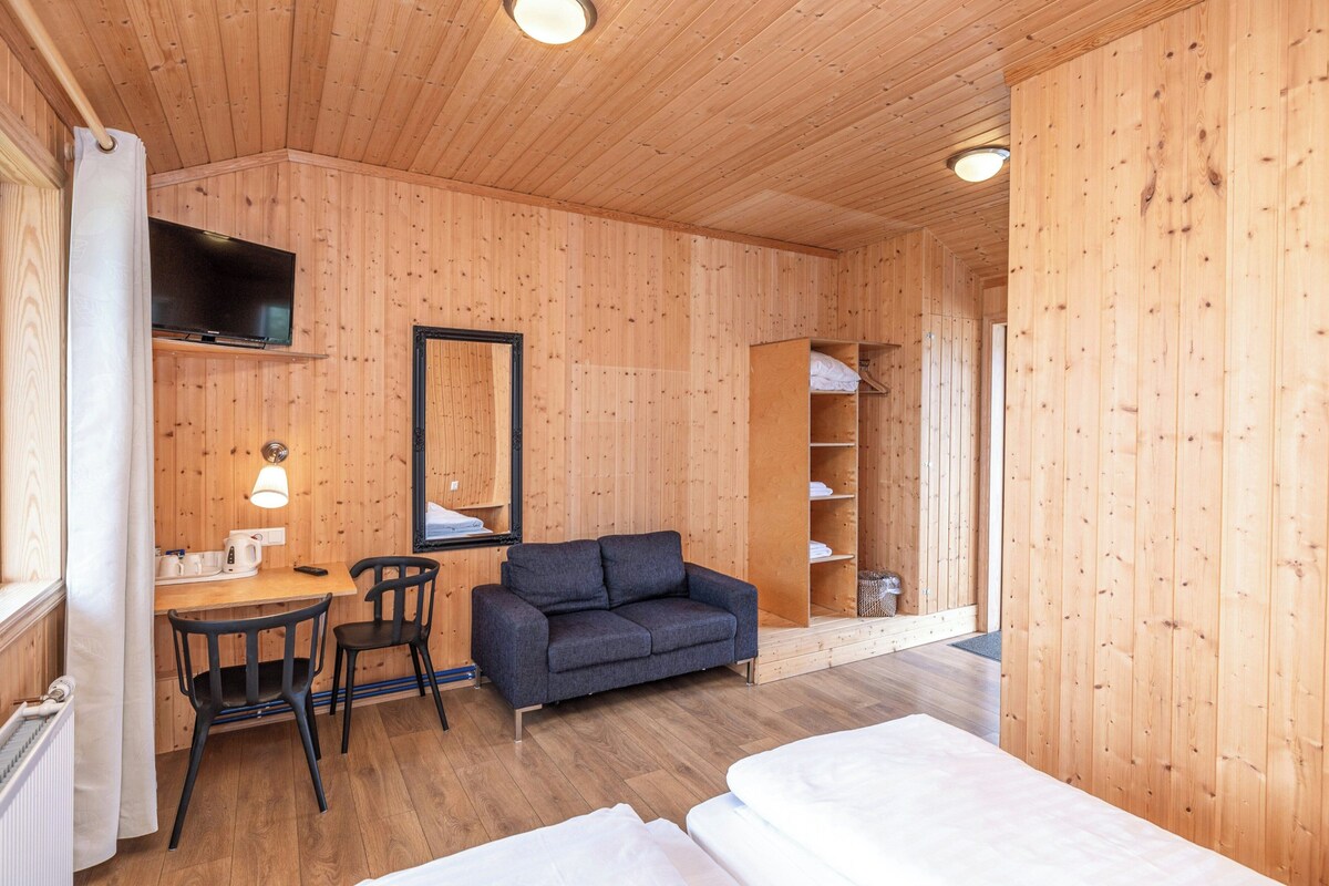 Quarto com painéis de madeira, um sofá, televisão, escrivaninha com abajur e uma cama.