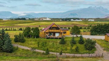 Uma casa de madeira de dois andares com varanda, emoldurada por uma paisagem rural com montanhas ao fundo.