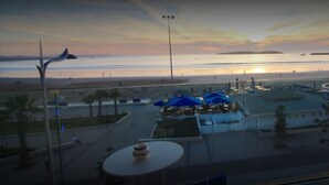 Vista de uma área à beira-mar com pôr do sol, um estacionamento e um prédio com toldos azuis.