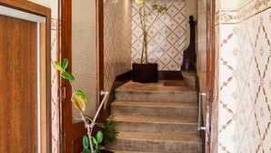Uma escadaria com um arco de madeira que dá acesso a um quarto com paredes estampadas e um vaso de planta.