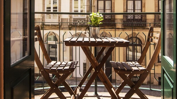 Uma varanda com mesa e cadeiras de madeira, um vaso de planta e vista para um edifício com janelas em arco e telhado telhado.