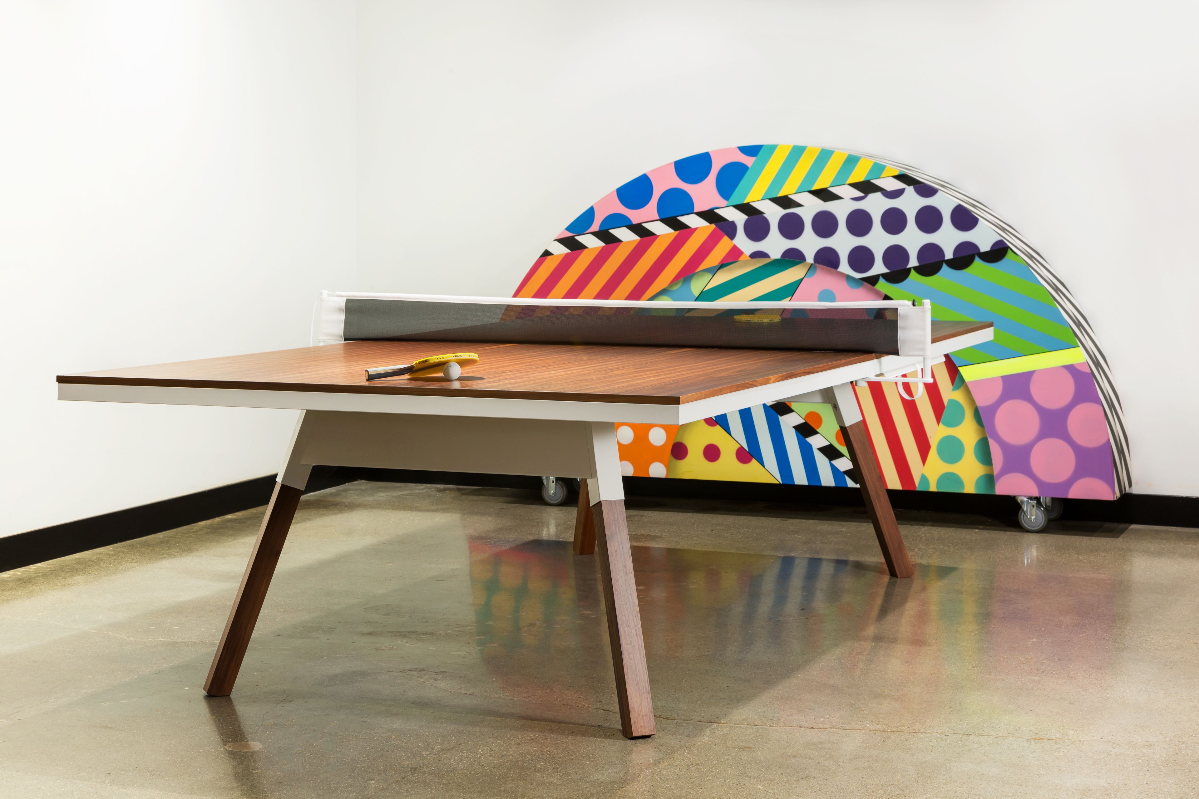 Uma mesa com tampo de madeira e pés de metal, posicionada em frente a uma parede colorida com bolinhas.