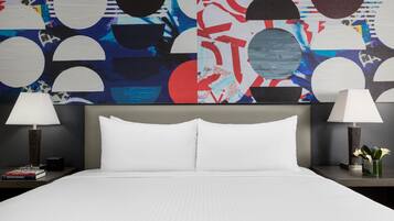 Quarto de hotel com uma grande pintura abstrata na parede, cama bem arrumada com roupa de cama branca, duas mesas de cabeceira com luminárias e livros, e um vaso com flores em uma das mesas.