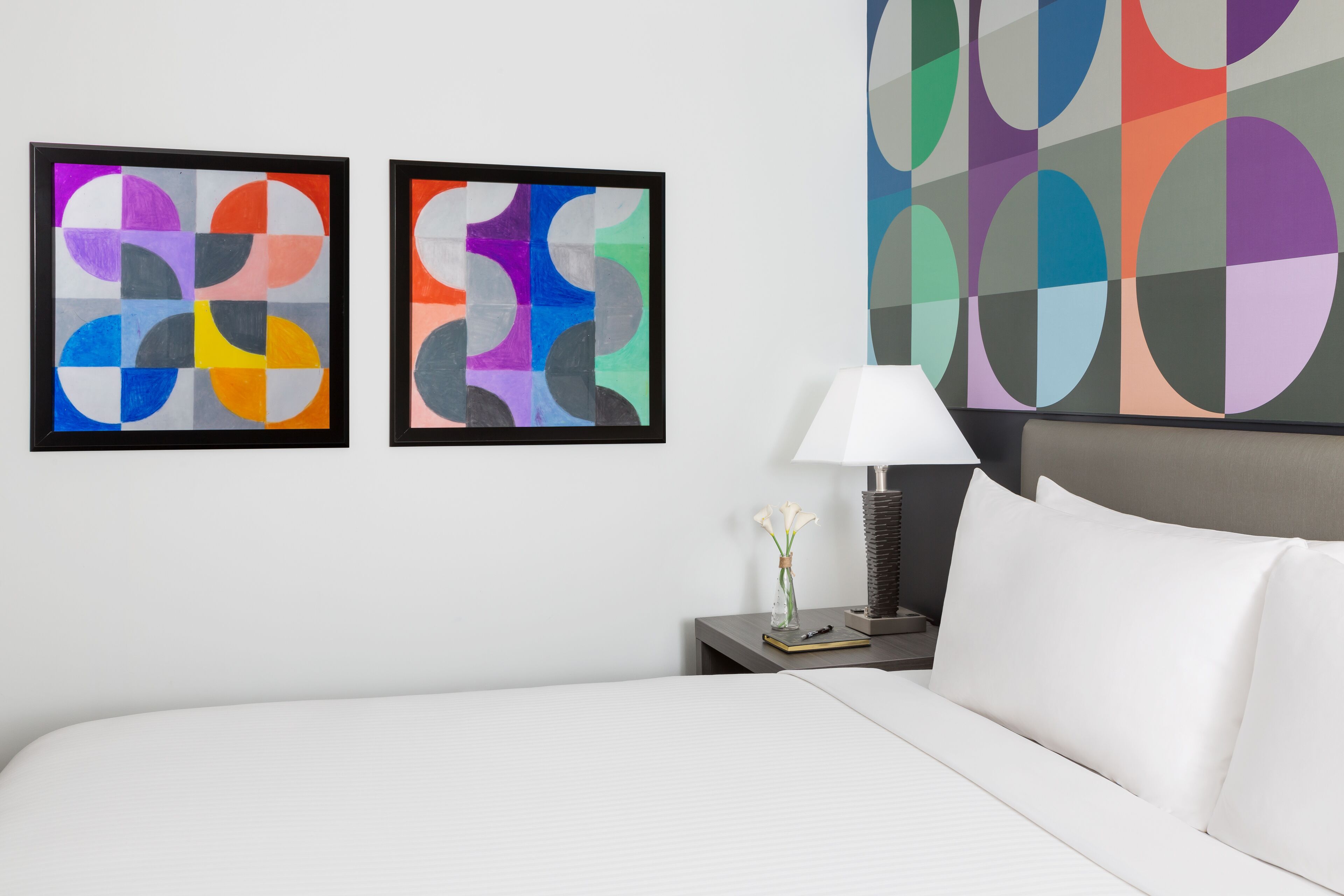 Quarto de hotel com uma cama bem arrumada, mesa de cabeceira com abajur e duas obras de arte abstratas nas paredes.