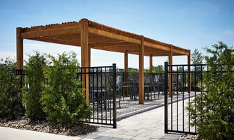 Estrutura de pérgola de madeira com cadeiras e mesas de metal por baixo, cercada por uma cerca de metal e vegetação.