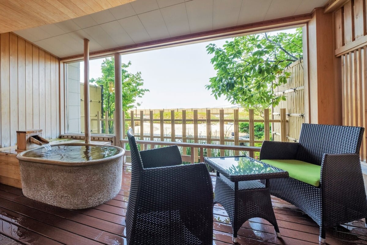 Um deck de madeira com hidromassagem, móveis de vime e vista para árvores e um cercado.