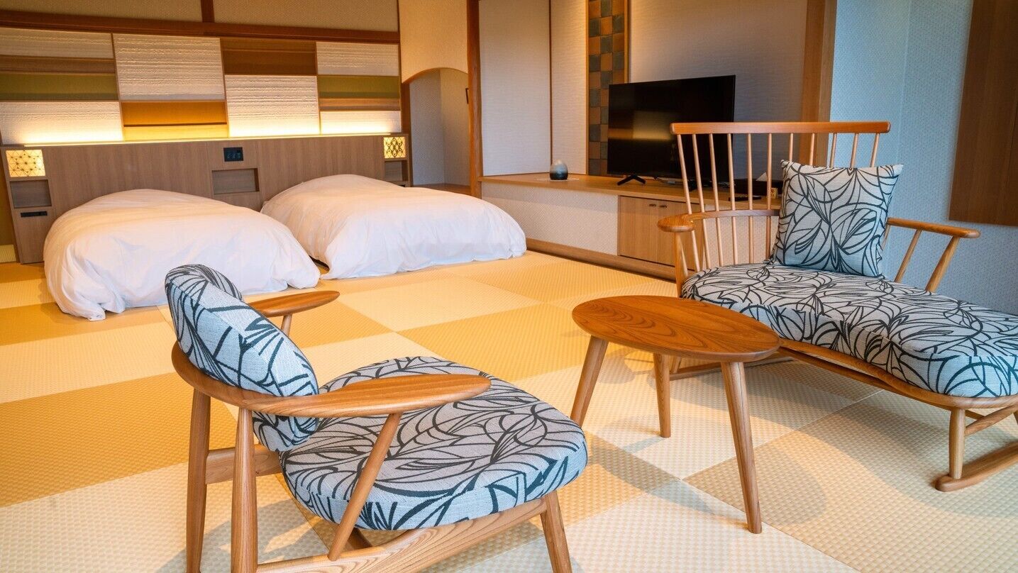 Quarto de hotel com uma cama, duas cadeiras, uma mesinha, uma televisão e uma parede com um padrão decorativo.