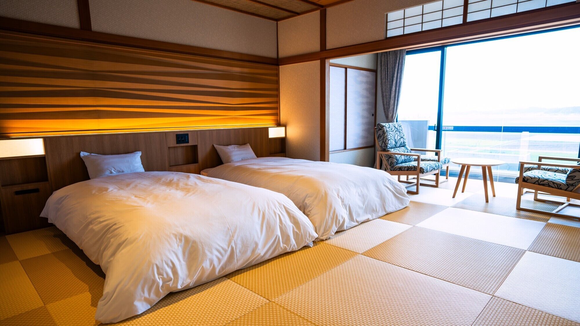 Quarto de hotel com duas camas, uma cadeira, uma mesa pequena e uma janela grande com vista.
